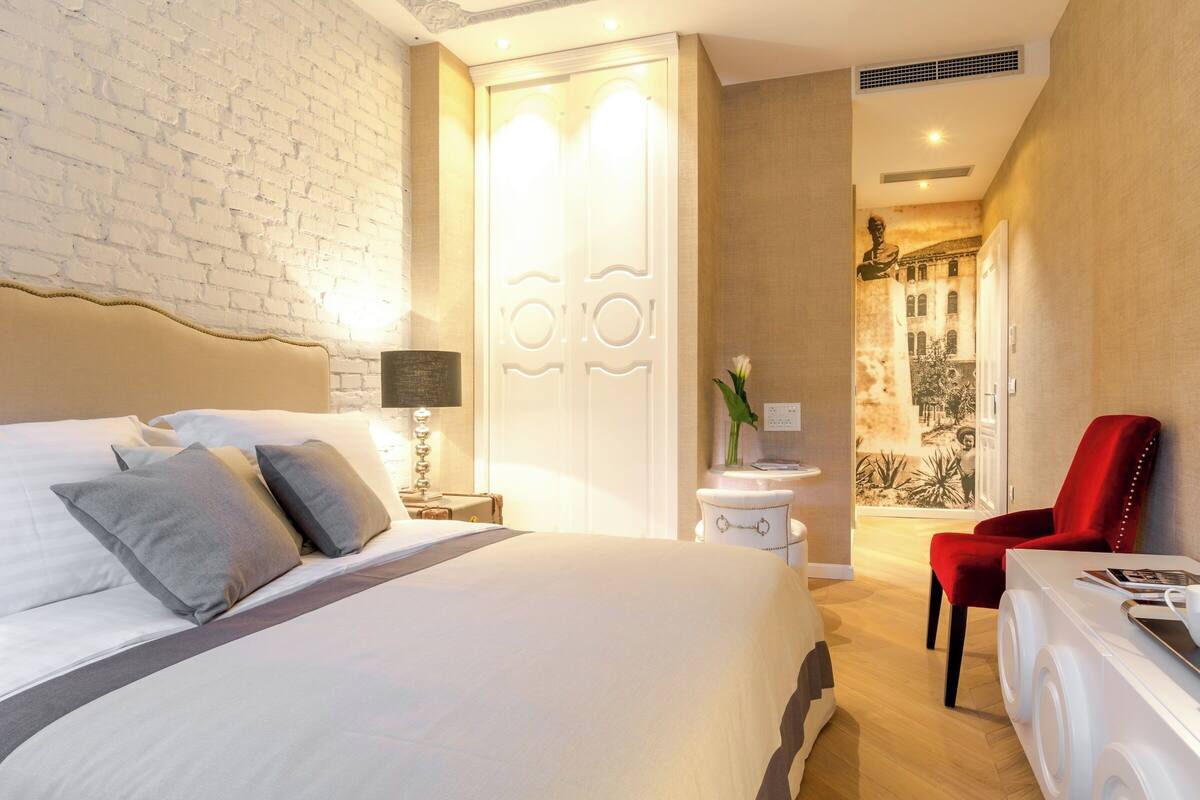 Quarto de hotel com uma cama, uma cadeira vermelha, uma mesinha e uma parede com padrão de tijolos.