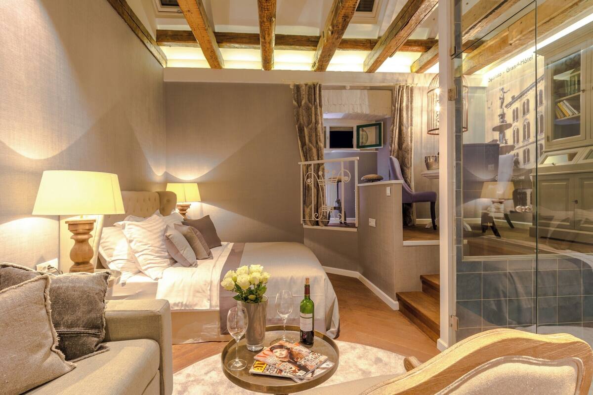 Quarto de hotel com cama, área de estar com sofá e mesinha de centro, banheiro com paredes de vidro e vista para o exterior.