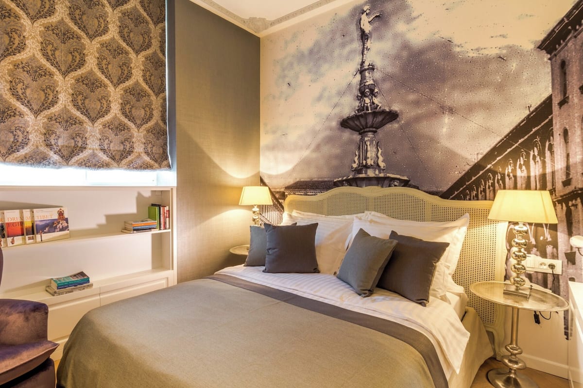 Quarto com cama, mesas de cabeceira, abajur, estante e um mural na parede.