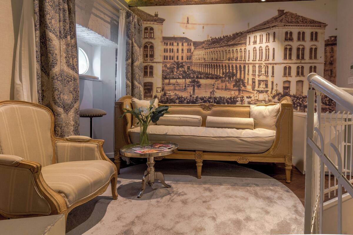 Quarto com sofá bege, duas poltronas, uma mesa redonda pequena com um vaso, e um mural grande de um edifício histórico.
