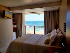 Quarto de hotel com cama, televisão, vista para a praia e varanda.