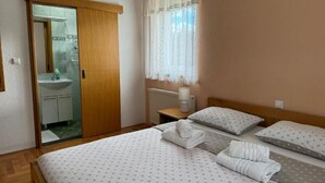 Quarto de hotel com porta de madeira que dá para um banheiro, uma cama com dois travesseiros e uma mesa de cabeceira com abajur.
