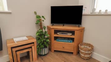 Quarto com televisão sobre um suporte de madeira, uma pequena mesa de madeira, uma planta em vaso, um jogo de Scrabble e uma cesta de palha.