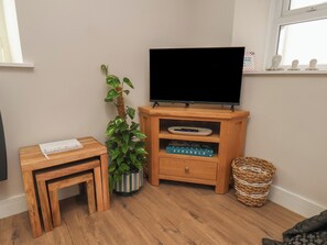 Quarto com televisão sobre um suporte de madeira, uma pequena mesa de madeira, uma planta em vaso, um jogo de Scrabble e uma cesta de palha.
