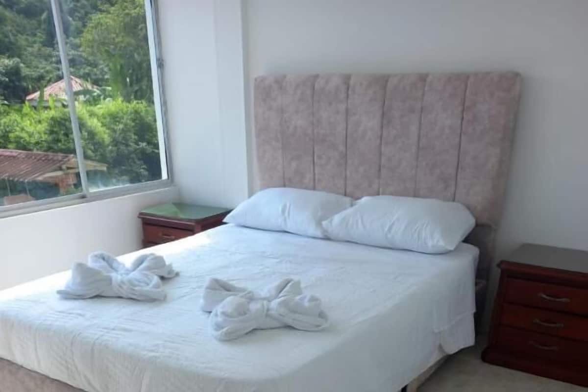 Cama bem arrumada com lençóis e toalhas brancos, uma janela grande com vista para o verde e uma mesa de cabeceira de madeira com gaveta.