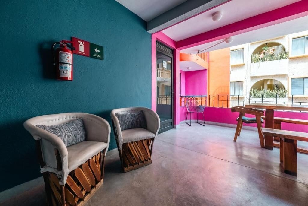 Quarto com paredes verde-água, um extintor de incêndio, duas poltronas estofadas, um banco de madeira e uma varanda com guarda-corrimão rosa.