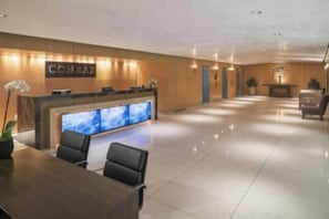 Uma área de recepção de hotel, com balcão, cadeiras e uma tela grande.