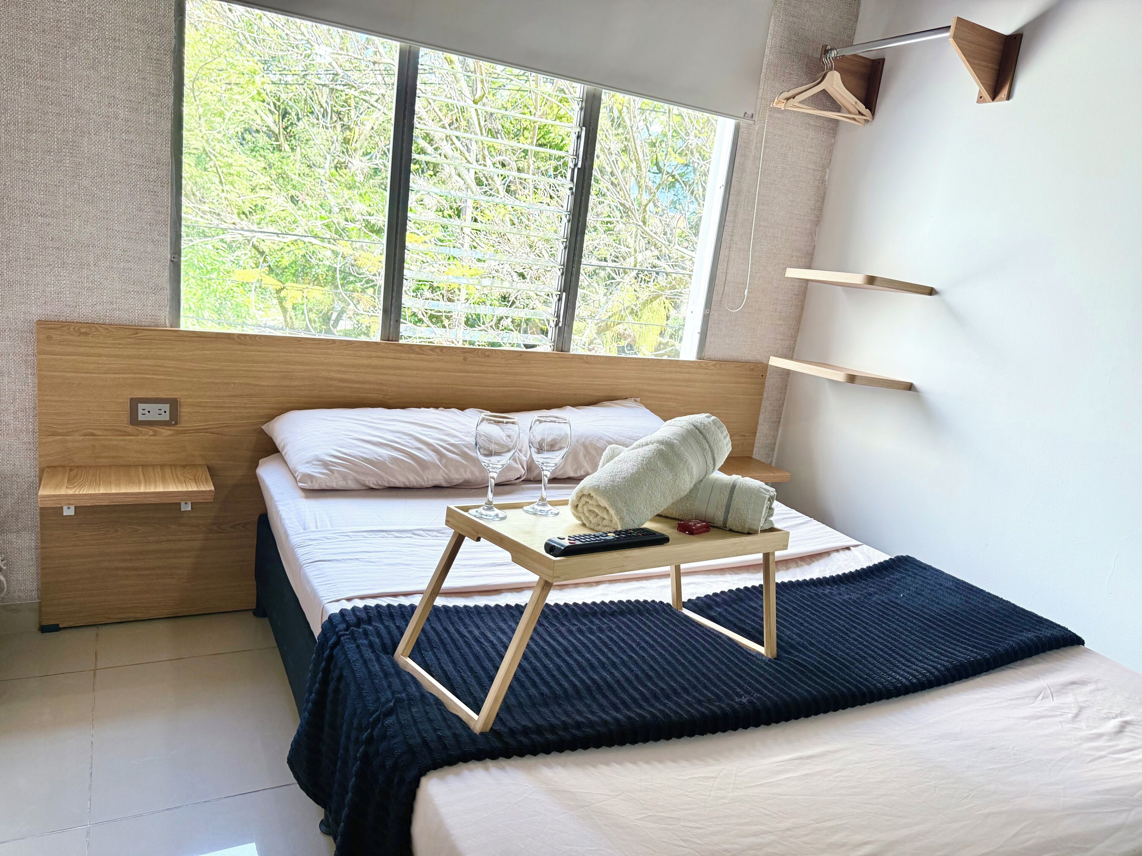 Quarto moderno com cabeceira de madeira, cama com roupa de cama branca, mesa de cabeceira com bandeja contendo copos e toalhas, e vista para o verde pela janela.