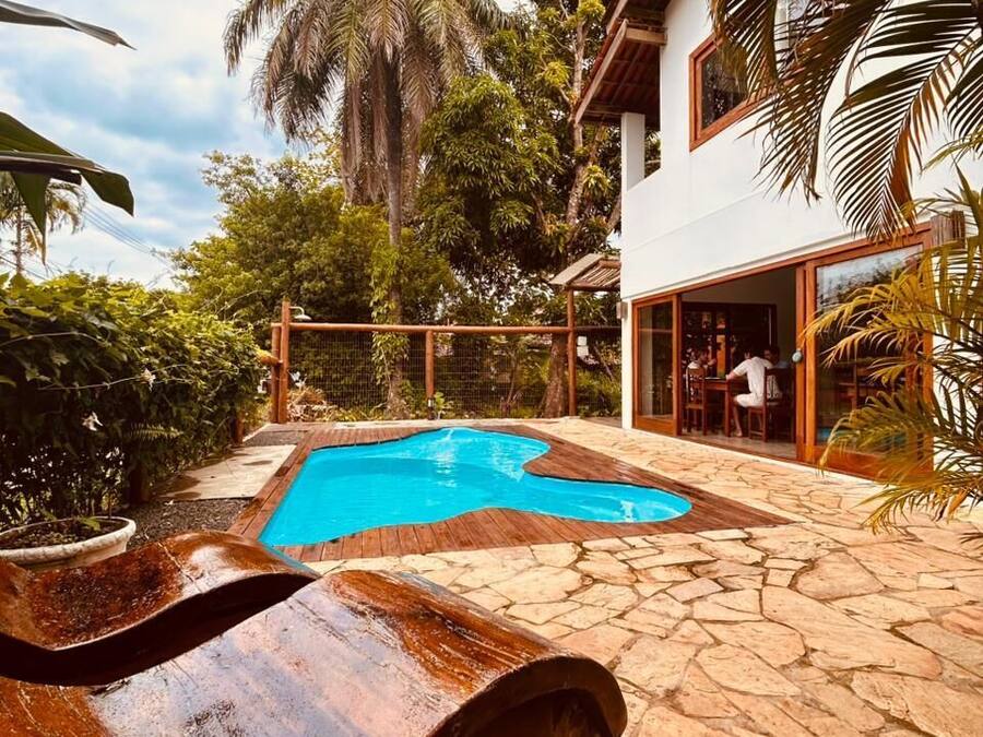 Uma piscina com deck de madeira, cercada por vegetação tropical e uma casa com grandes janelas.