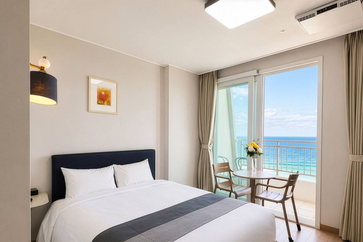 Quarto de hotel com cama, mesa de cabeceira, mesa com cadeiras, varanda com vista para o oceano e um quadro na parede.