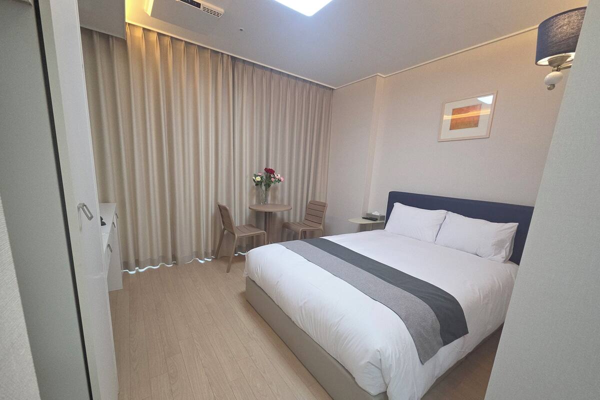 Quarto de hotel com cama, escrivaninha, cadeira, mesa pequena e cortina.