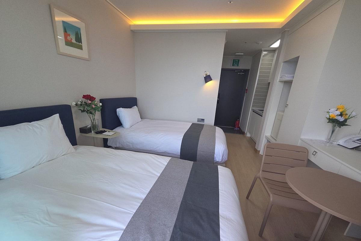 Quarto de hotel com duas camas, uma mesa pequena, uma cadeira, um vaso com flores e um quadro na parede.