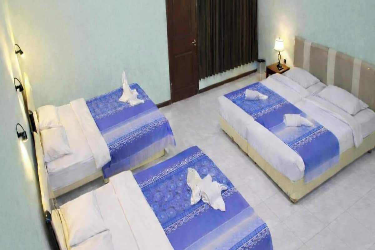 Quarto de hotel com duas camas, uma porta, uma janela com cortinas e uma mesinha de cabeceira com abajur.