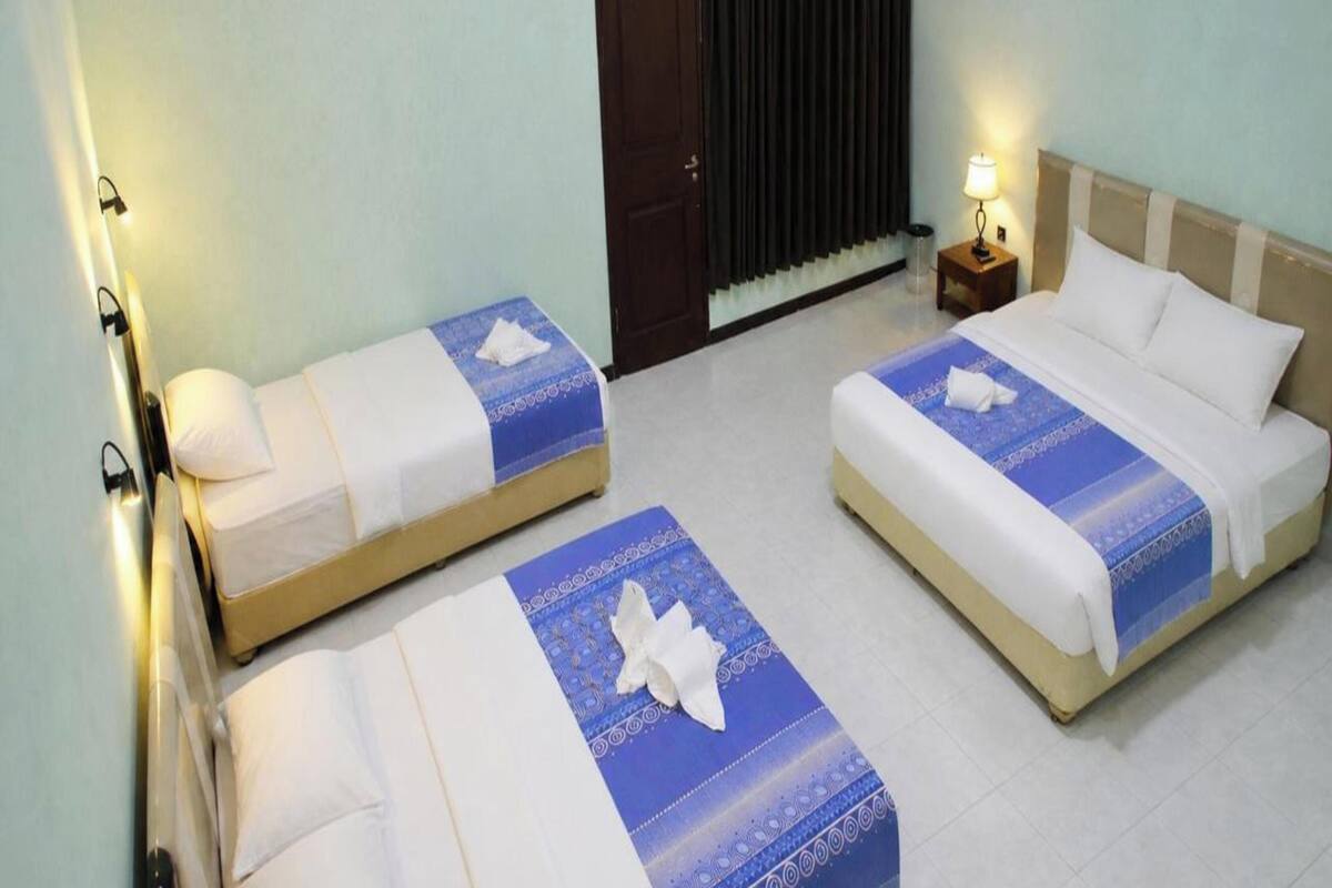 Quarto de hotel com duas camas, uma porta, janela com cortinas, mesa de cabeceira com abajur e luminária na parede.