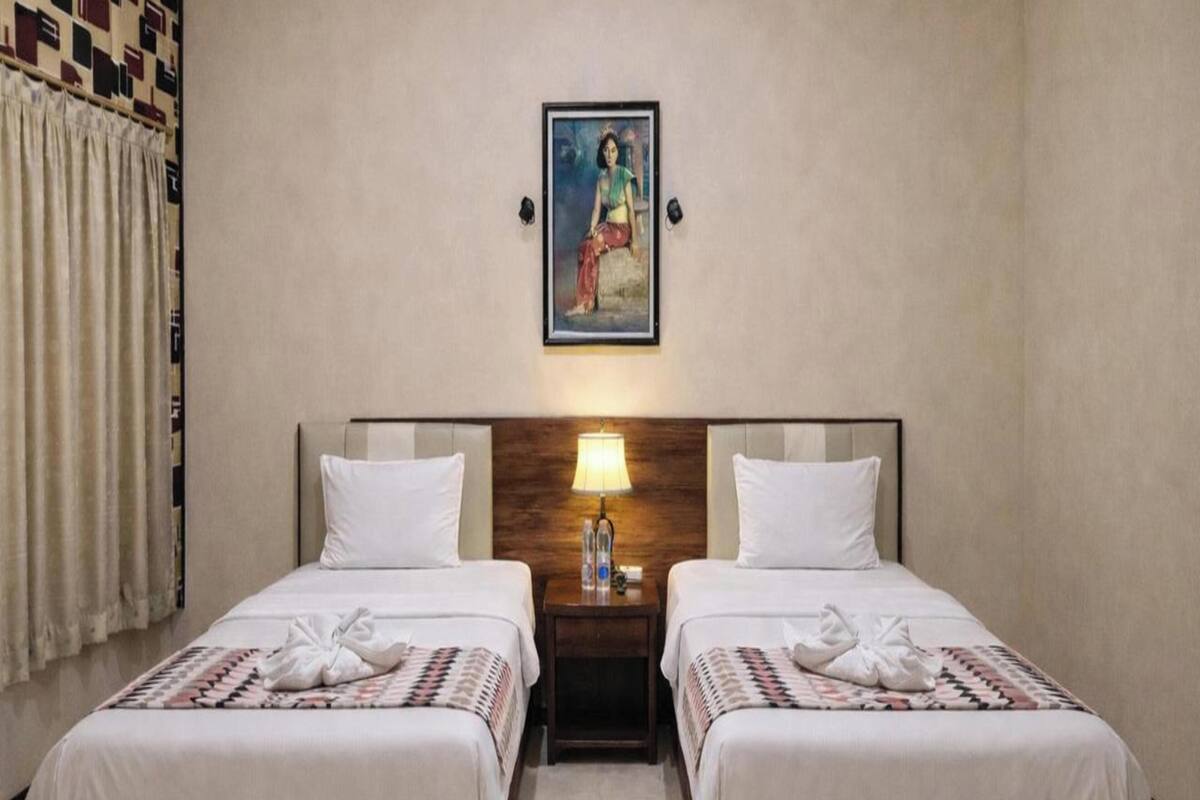 Quarto de hotel com duas camas, cabeceira com luminária, pintura na parede e cortina estampada.
