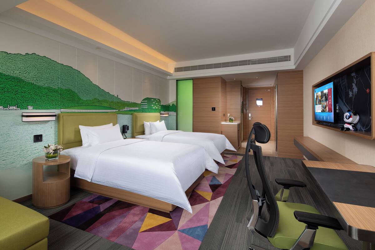 Quarto de hotel com cama, duas mesas de cabeceira, escrivaninha com cadeira, televisão e um mural na parede.