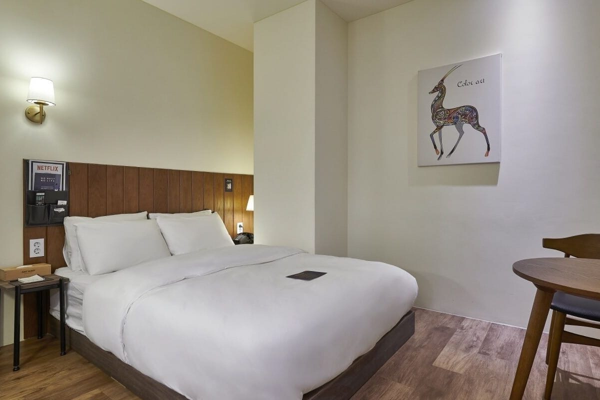 Quarto de hotel com cama, mesa de cabeceira, luminária, obra de arte na parede e uma pequena mesa com uma cadeira.