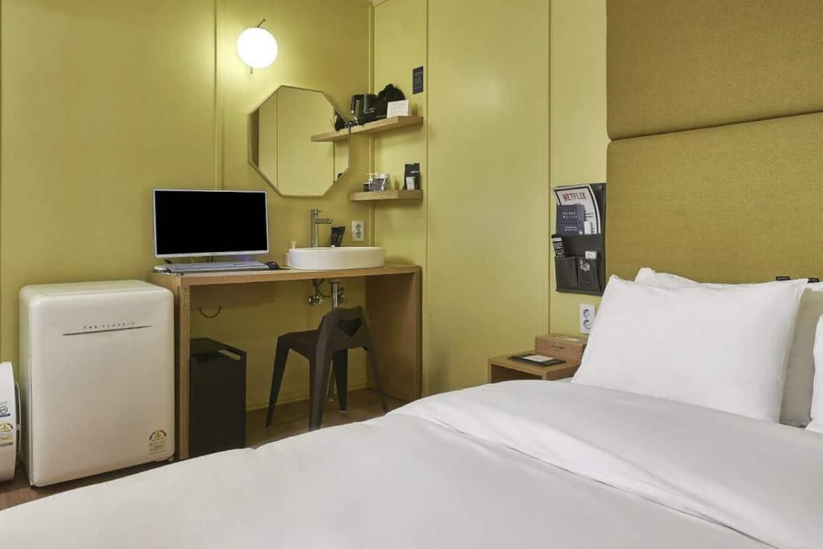 Quarto de hotel com uma cama, uma secretária com um computador, um frigorífico pequeno, um espelho e uma prateleira com artigos de higiene pessoal.