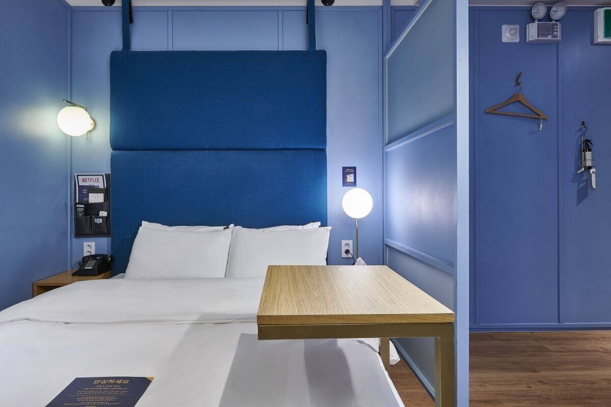 Quarto de hotel com cama, mesa de cabeceira, unidades de ar condicionado fixadas na parede, cabides e um aviso indicando serviço Netflix.