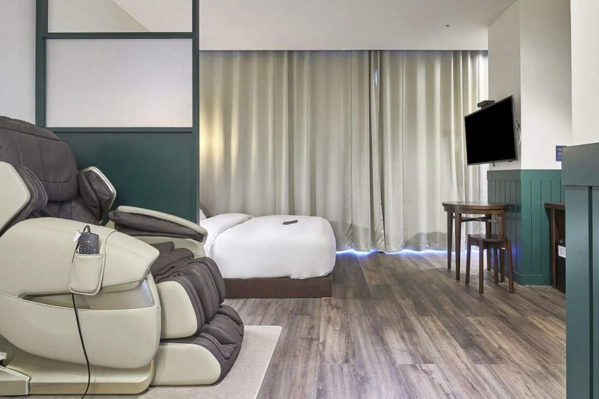 Quarto de hotel com cadeira de massagem, cama, televisão e uma mesinha.