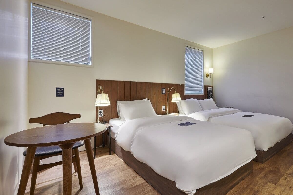 Quarto de hotel com duas camas, uma cabeceira de madeira, uma mesinha e duas luminárias de cabeceira.