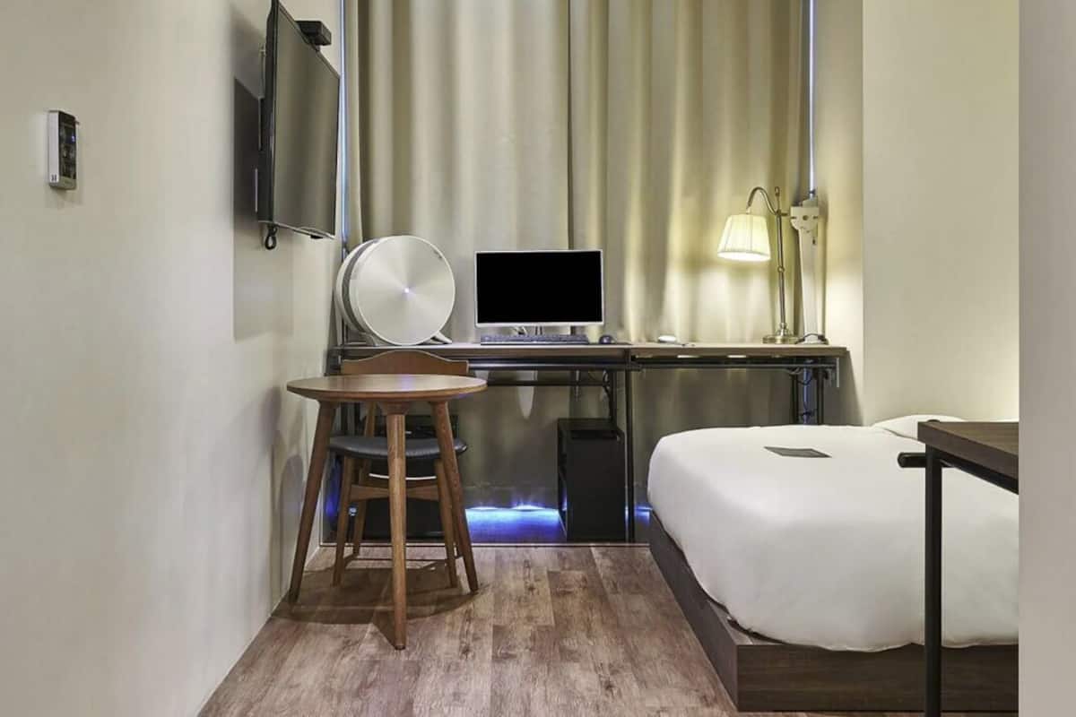 Quarto de hotel com uma cama, escrivaninha com computador, cadeira, televisão e luminária.