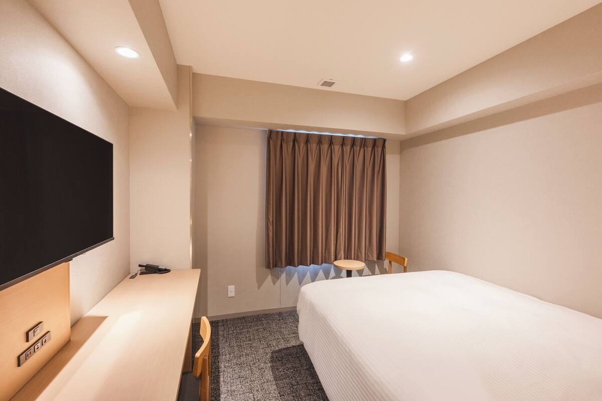 Quarto de hotel com cama, televisão de tela plana, escrivaninha, cadeira, mesinha e janela com cortinas.