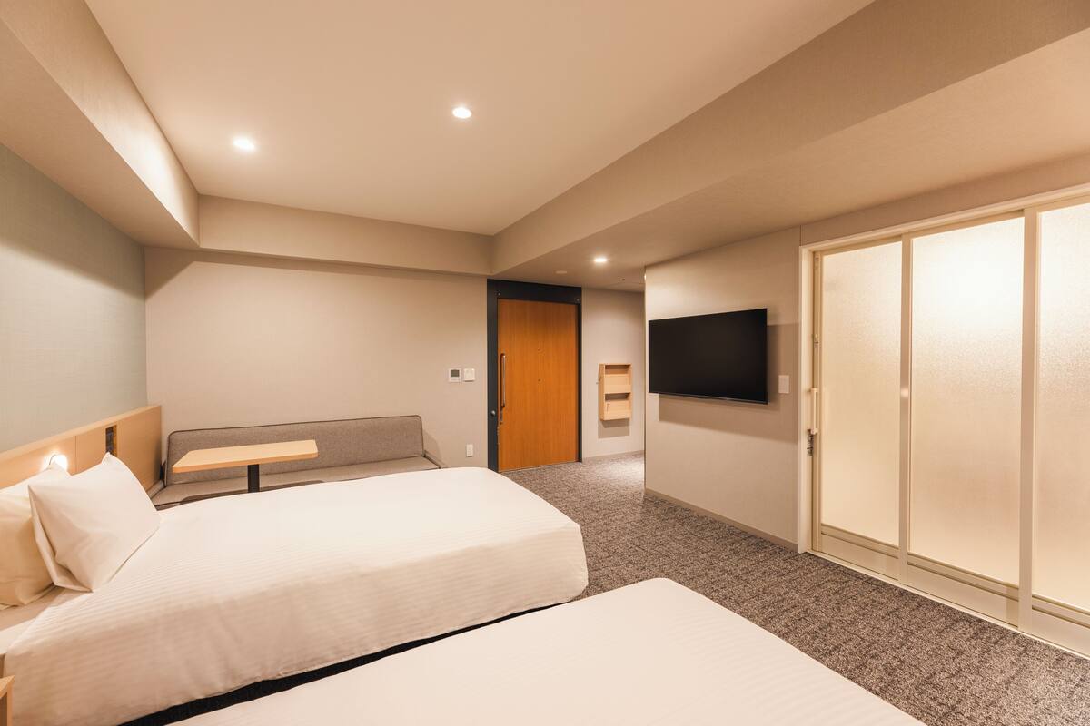 Quarto de hotel com duas camas, uma televisão, uma porta de correr e um banco.