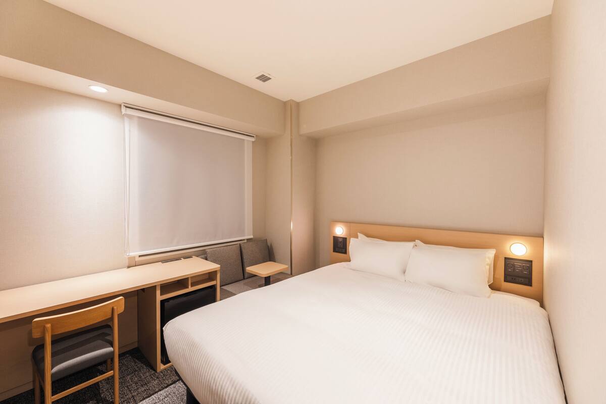 Quarto de hotel com cama, escrivaninha, cadeira, mesinha e luminária na parede.