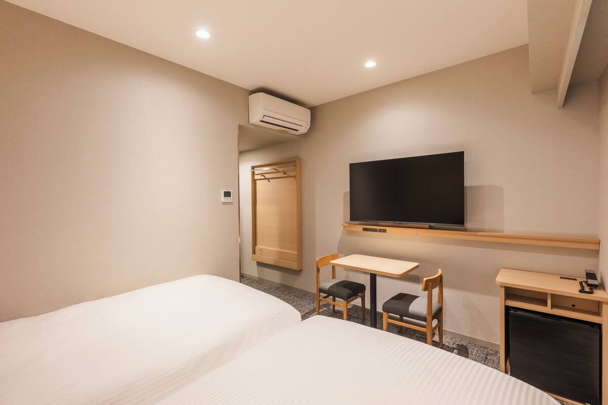 Quarto de hotel com duas camas, televisão na parede, secretária com cadeira e um pequeno frigorífico.
