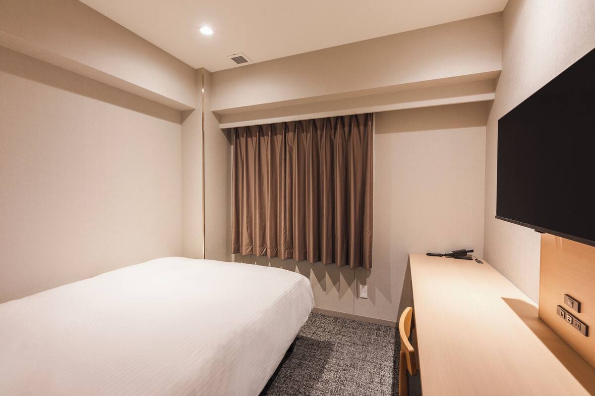 Quarto de hotel com cama, escrivaninha, televisão e janela com cortinas.