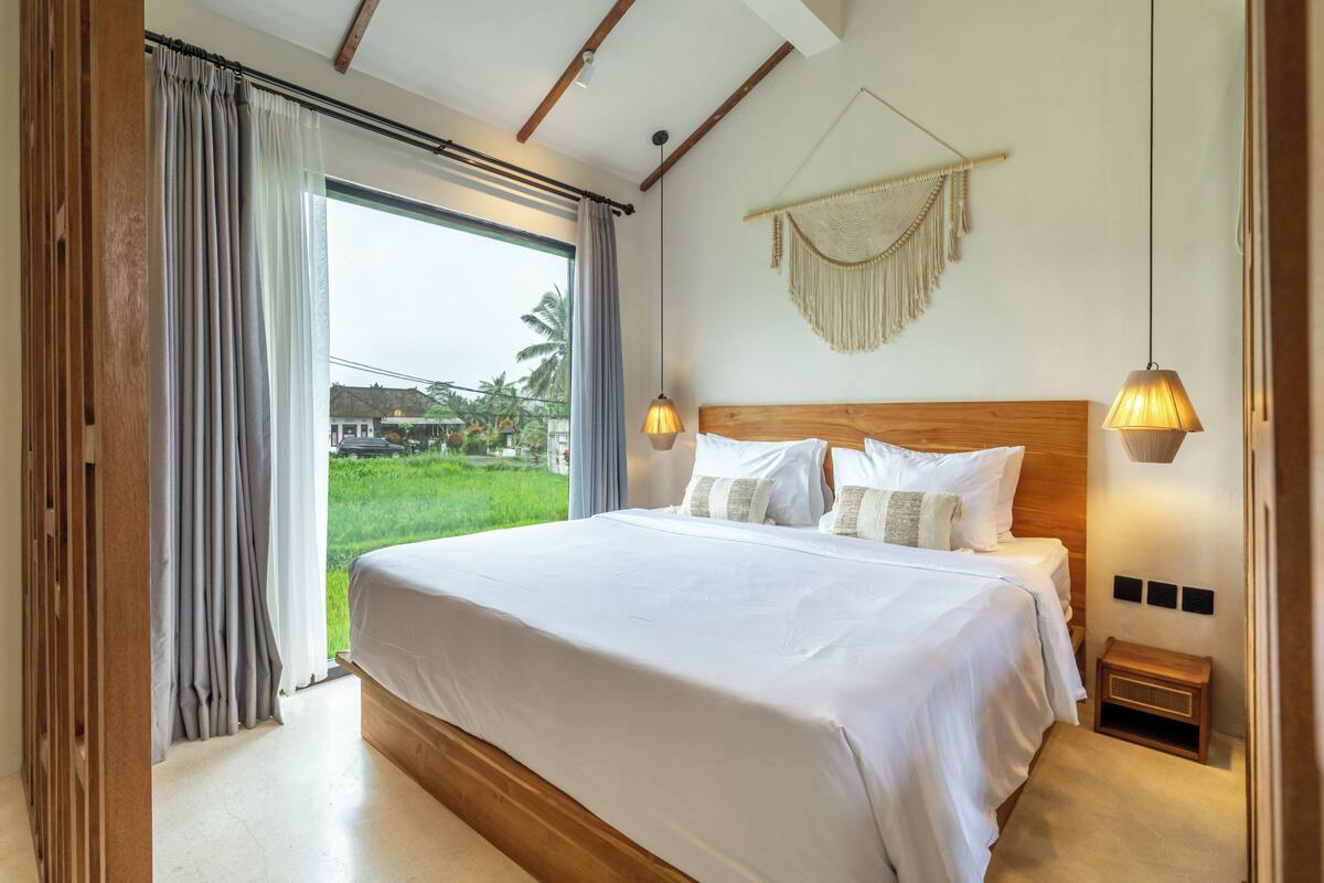 Quarto com uma cama grande, cortinas, vista para o verde, um pendente de macramé na parede e um abajur.