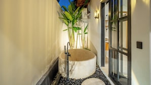 Uma banheira de pedra circular em um espaço estreito, com teto de madeira e uma planta grande.