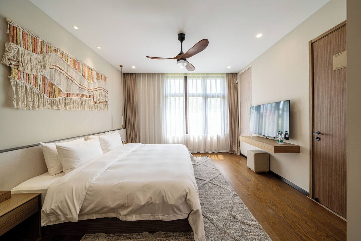 Quarto de hotel moderno com uma cama grande, ventoinha no teto, televisão de tela plana e uma janela ampla com cortinas.