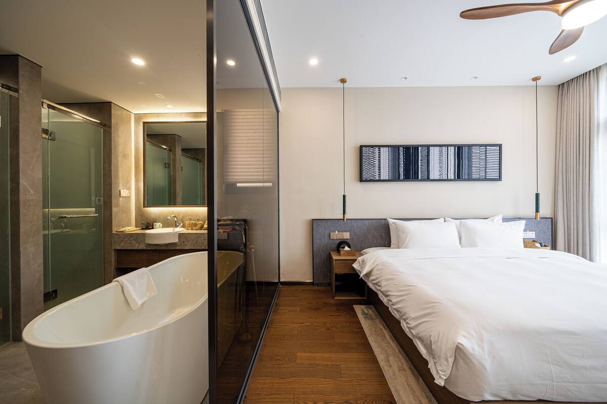 Quarto de hotel moderno com uma banheira grande, área de chuveiro separada, cama com lençóis brancos, mesa de cabeceira e uma obra de arte na parede.