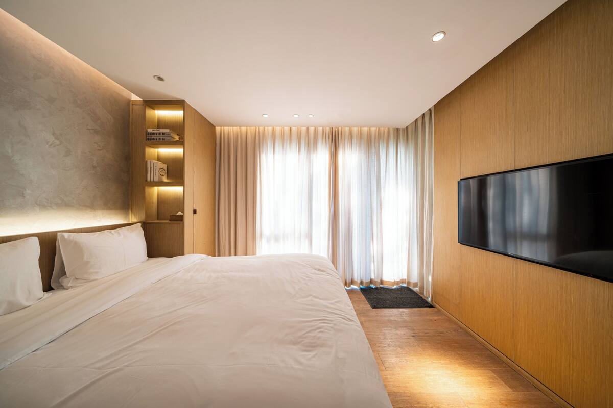 Quarto de hotel moderno com uma cama grande, televisão de tela plana e uma estante embutida com livros.
