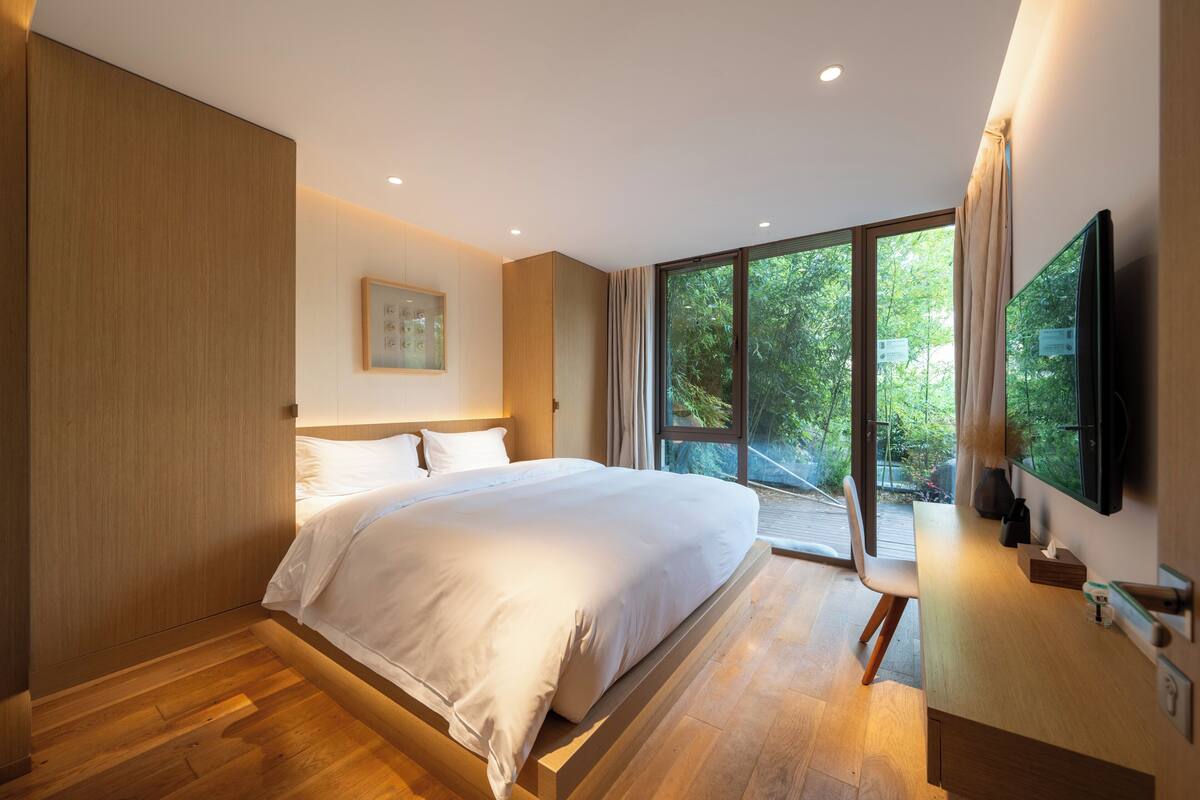 Quarto moderno com cama grande, mesa de cabeceira, televisão e janela com vista para o verde.