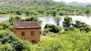 Um pequeno edifício térreo com telhado de azulejos, cercado por vegetação exuberante e um corpo d'água.