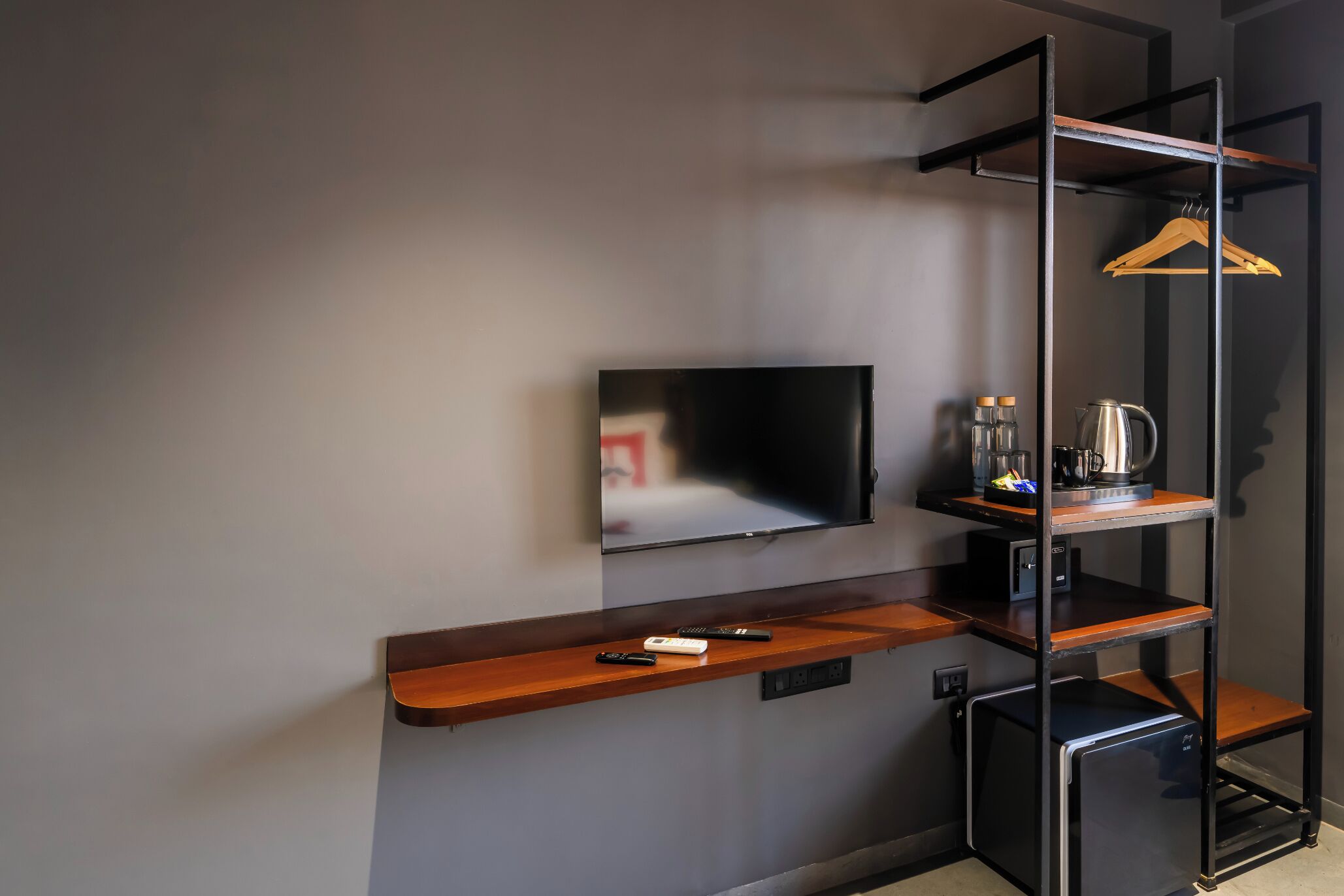 Quarto moderno com televisão de tela plana na parede, mesa de console de madeira com controle remoto, unidade de armazenamento preta com prateleiras e cabide de madeira pendurado.