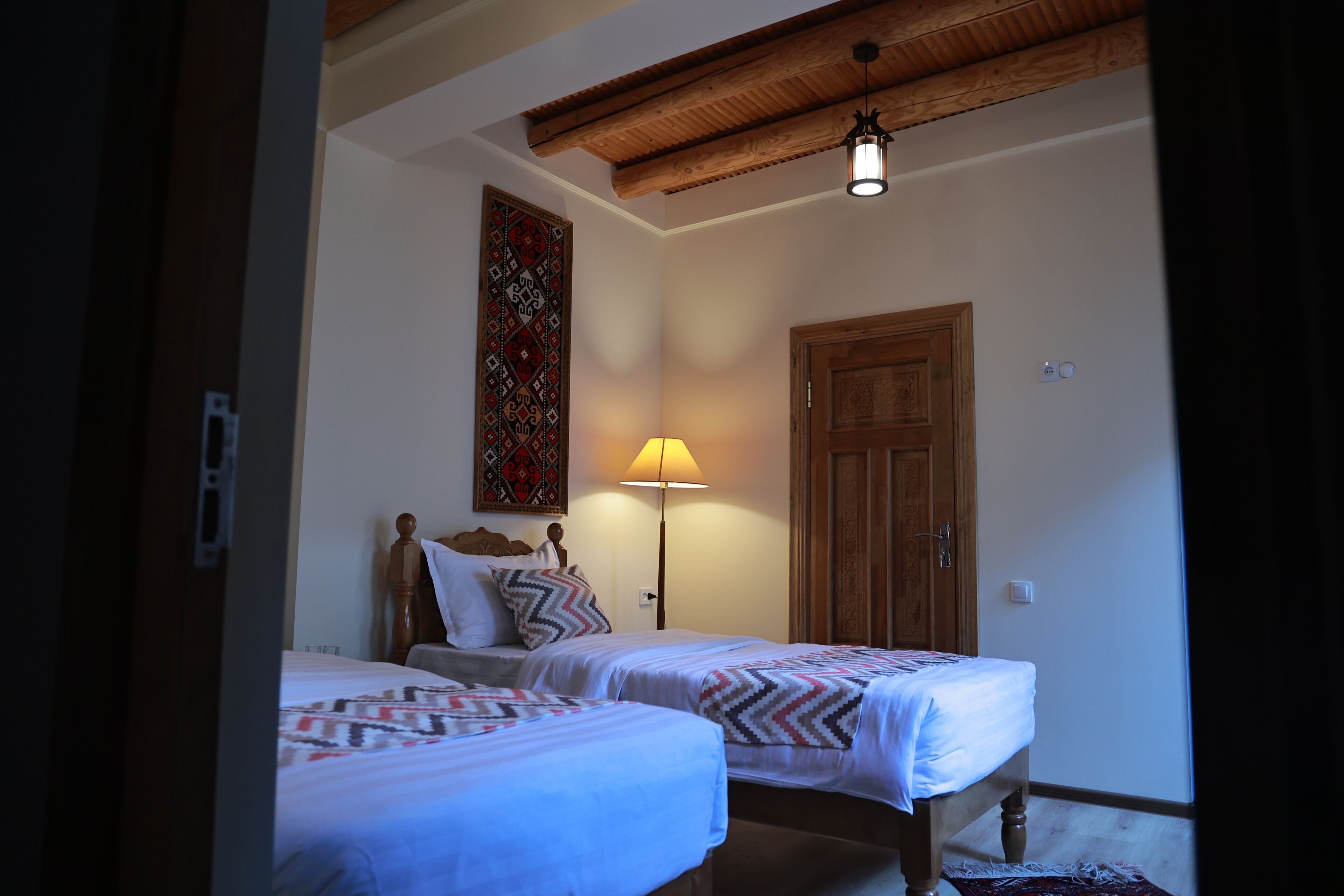 Quarto com duas camas, teto de madeira, luminária na parede e uma porta.