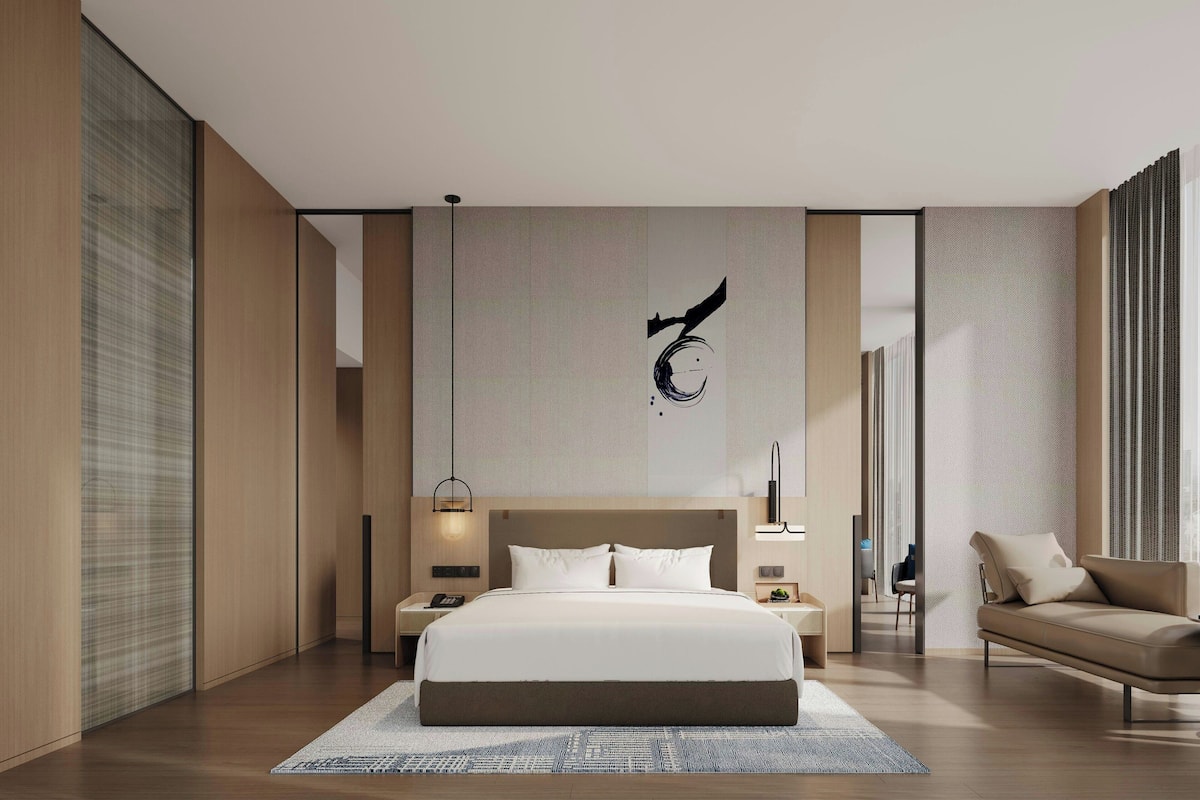 Quarto de hotel moderno com uma cama grande, um sofá, uma mesa de cabeceira e uma obra de arte na parede.