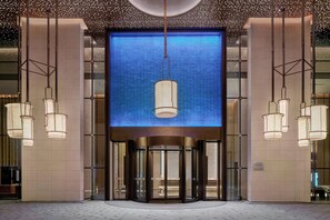 Entrada de hotel com porta giratória, uma tela iluminada grande e luminárias pendentes.