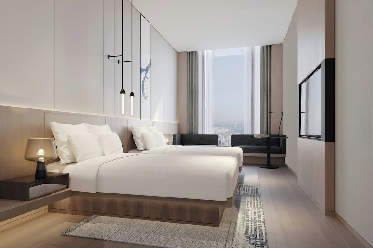 Quarto de hotel moderno com uma cama grande, uma mesa de cabeceira com abajur, TV fixada na parede e vista da cidade através de amplas janelas.