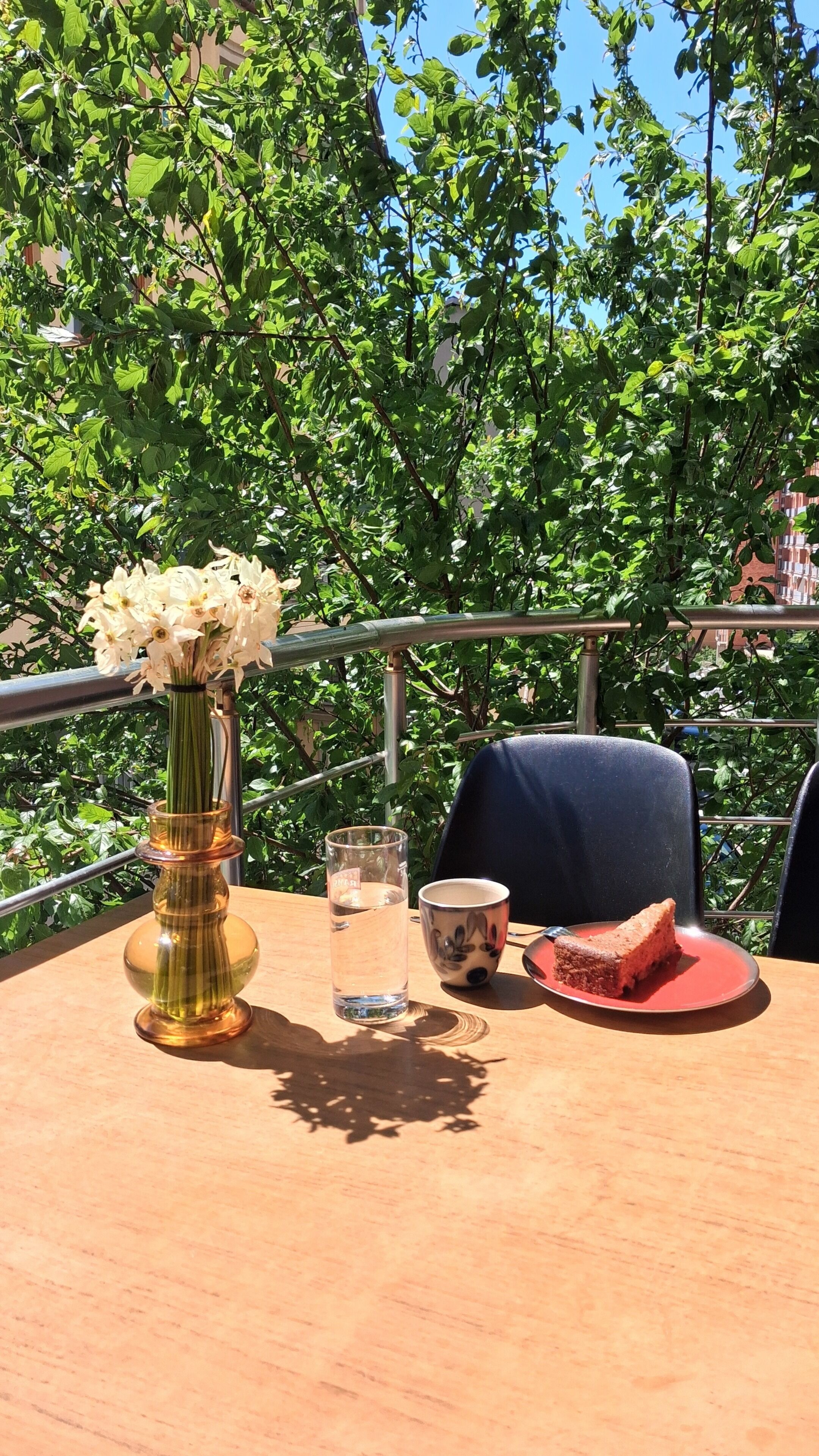 Uma mesa com um vaso de flores, um copo de água, uma caneca e uma fatia de bolo em um prato vermelho.