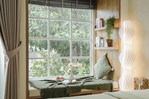 Quarto com uma janela grande, cama com manta verde, vaso branco com flores e luminária branca.