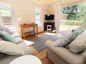 Interior acolhedor de uma cabana com sofá, televisão, lareira e um deck de madeira lá fora.