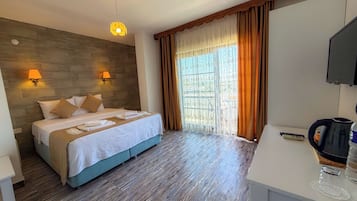 Quarto de hotel com cabeceira de madeira, cama com roupa de cama branca, duas luminárias fixadas na parede, uma televisão e uma grande janela com cortinas.
