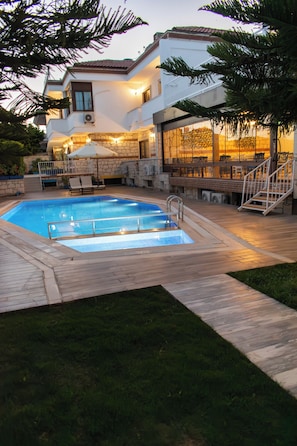 Piscina com deck de madeira, edifício branco com varanda e escadaria.