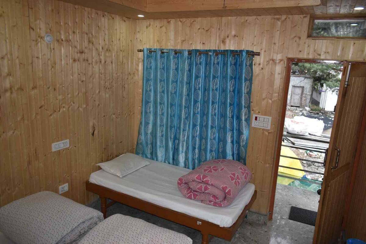 Um quarto pequeno de madeira, com uma cama, um banco e uma janela com cortinas.
