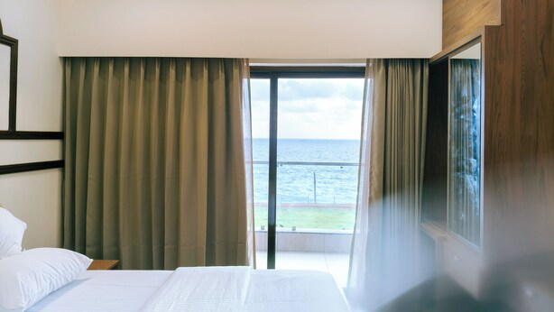 Quarto de hotel com cama, cortinas, televisão e vista para o mar.